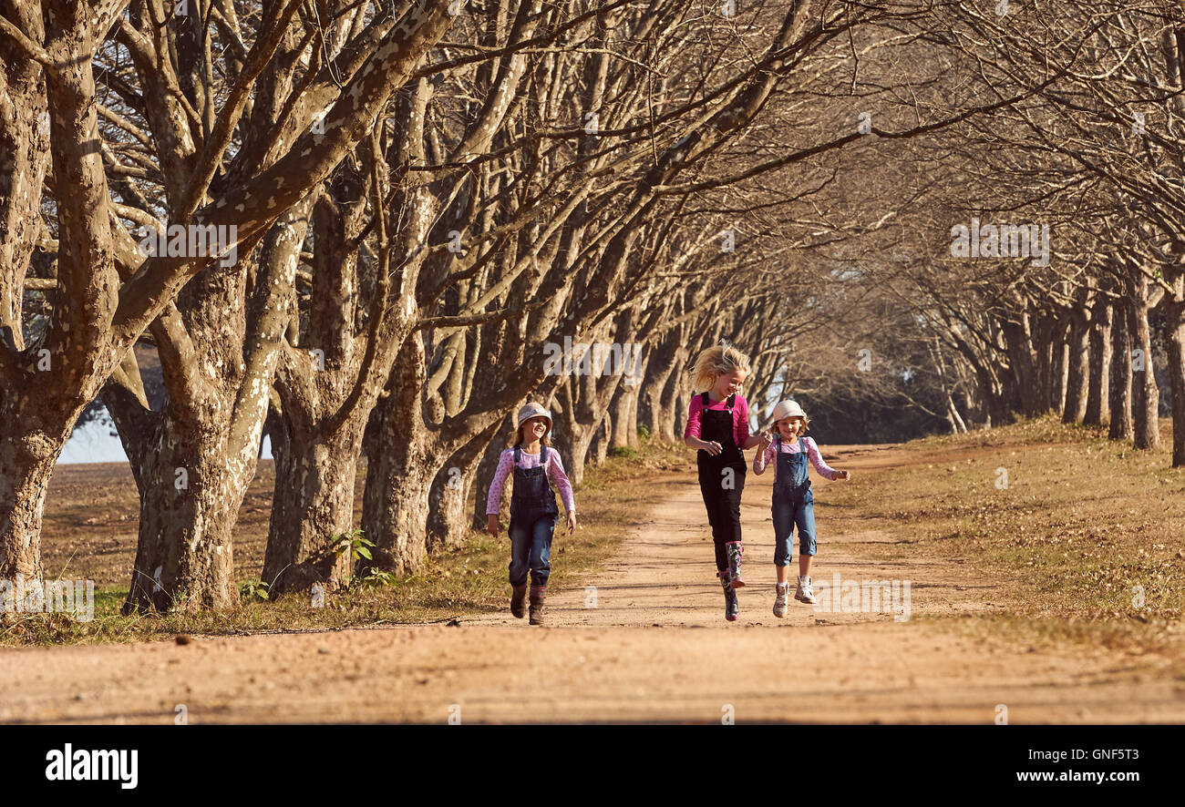 Three girls sisters running skipping down dirt road tree lined avenue ...