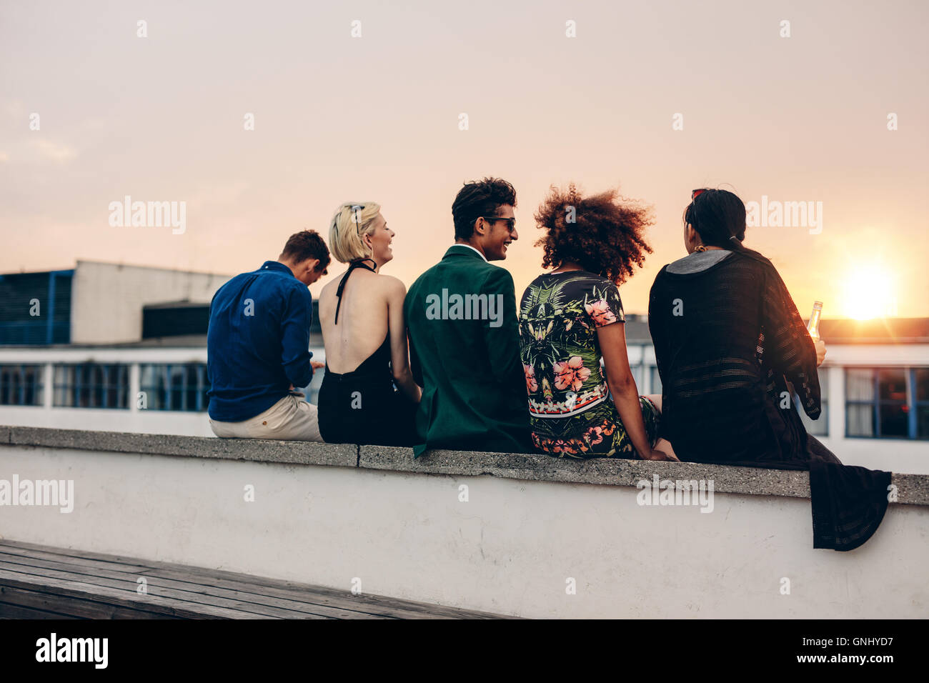 Rear view shot of young men and women sitting together on rooftop ...