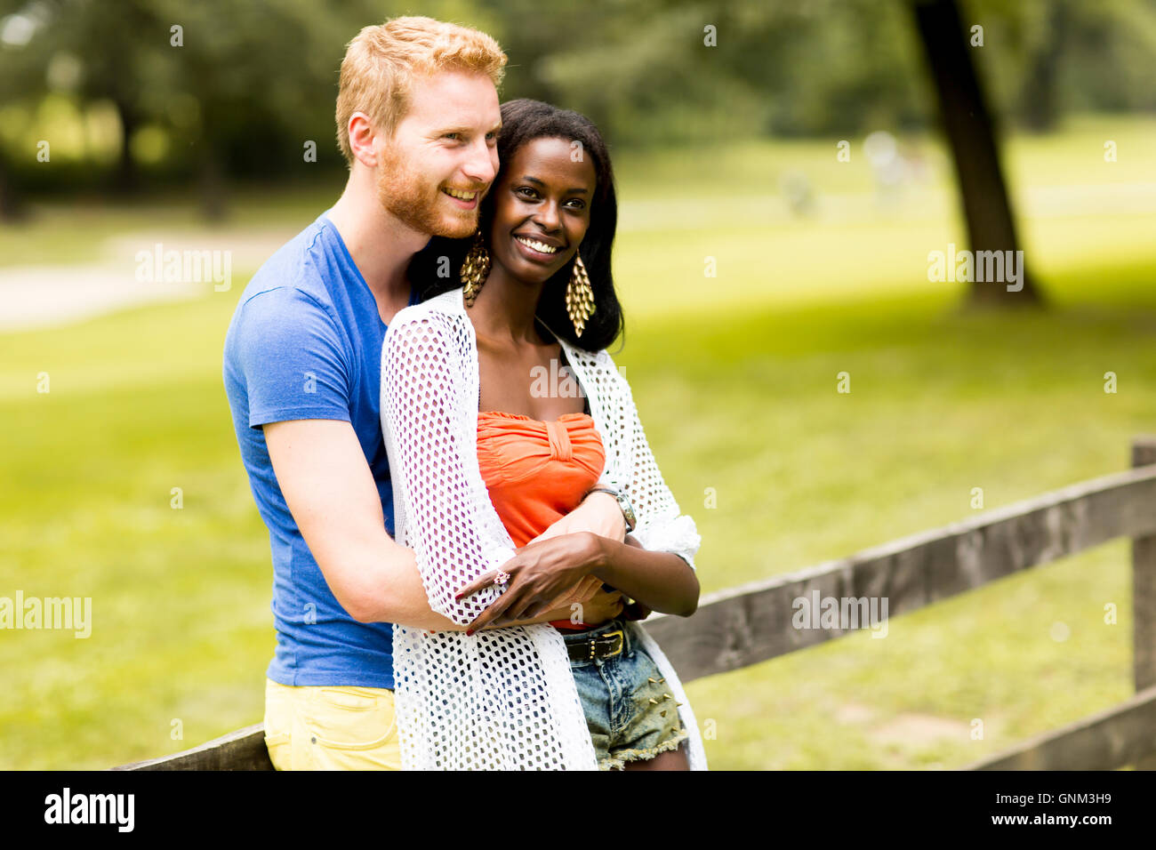 Young multiracial couple in love standing next to the fence in the park ...