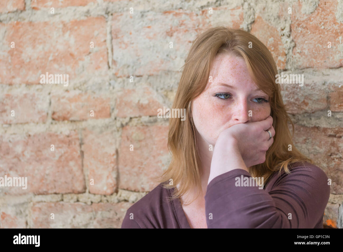 Portrait of sad redhead young woman is holding her face. Brick wall is ...