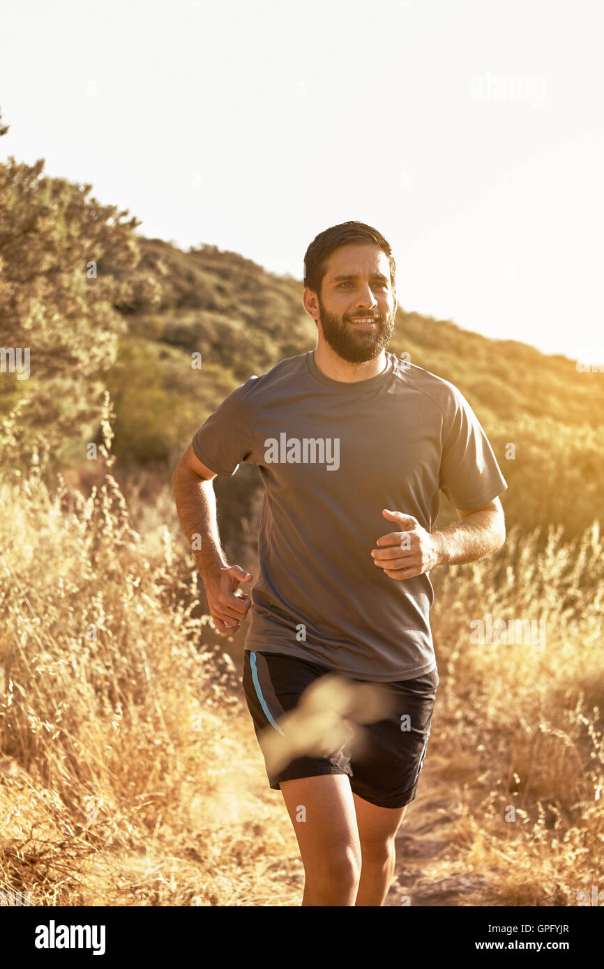 Young man jogging down a dusty path in full sun light with shrubs ...