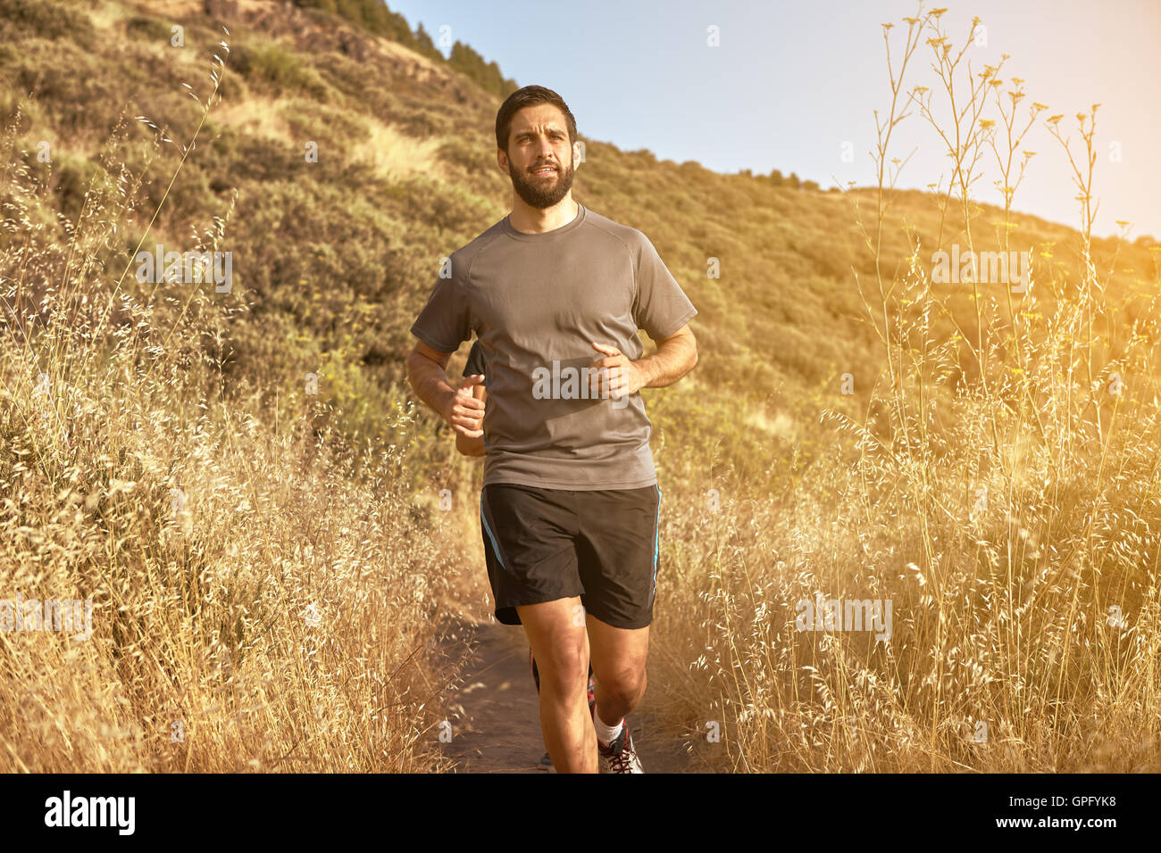 Young man jogging down a dusty path in full sun light with drying grass ...