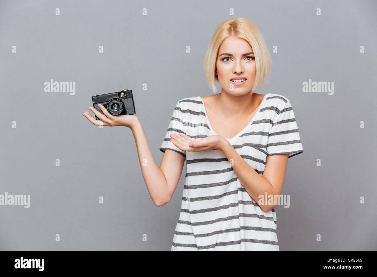 Confused blonde young woman holding old vintage photo camera over gray ...