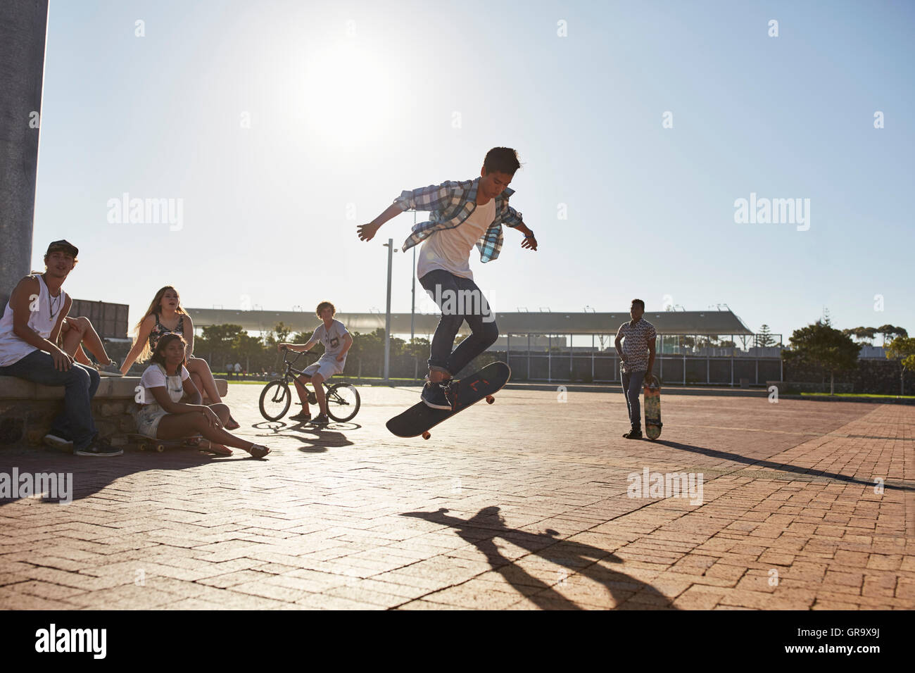 Friends watching teenage boy flipping skateboard at sunny skate park ...