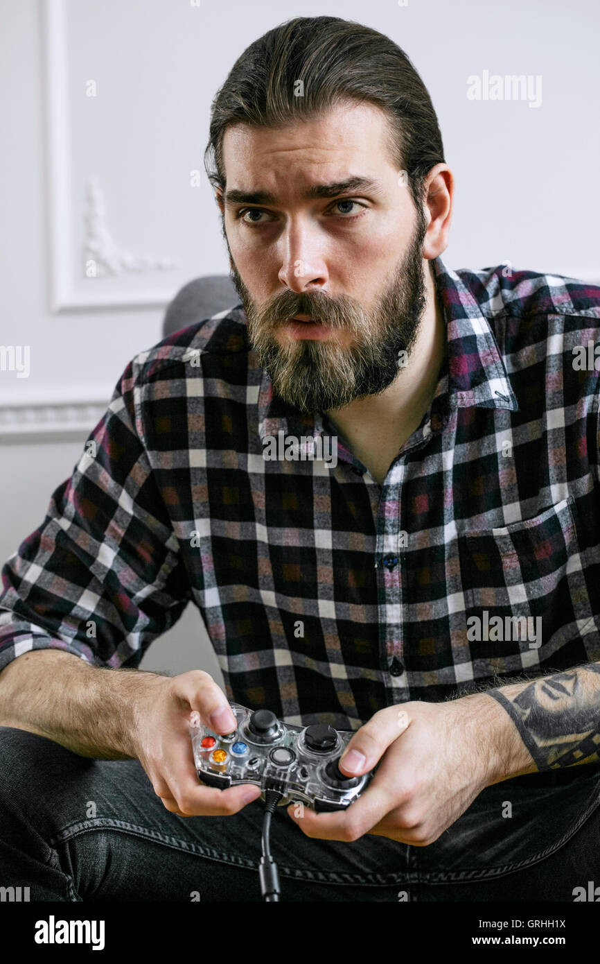 A young hipster with beard plays a video game seated with strength in ...