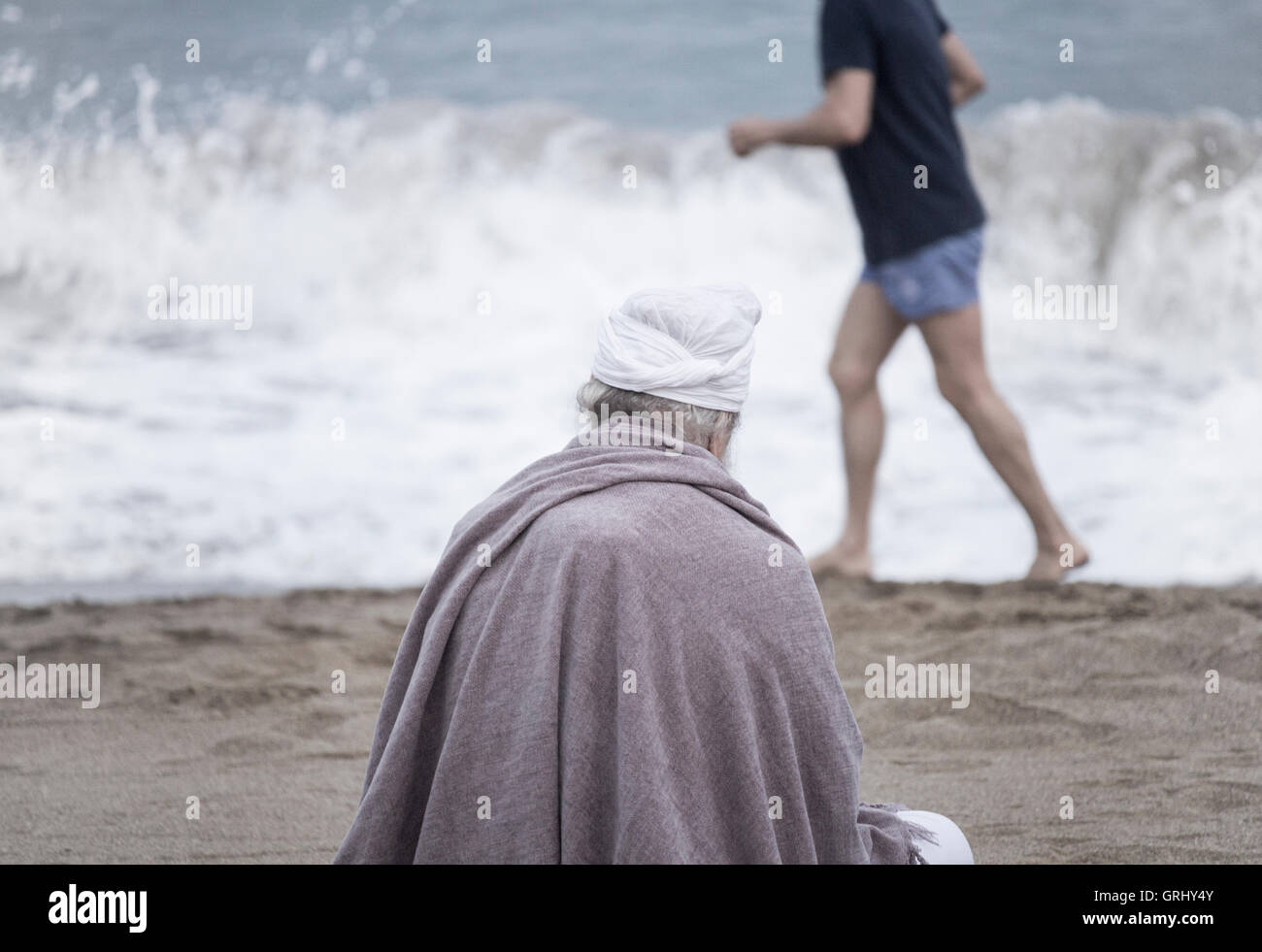 Elderly man wearing turban sitting on beach facing the ocean with ...