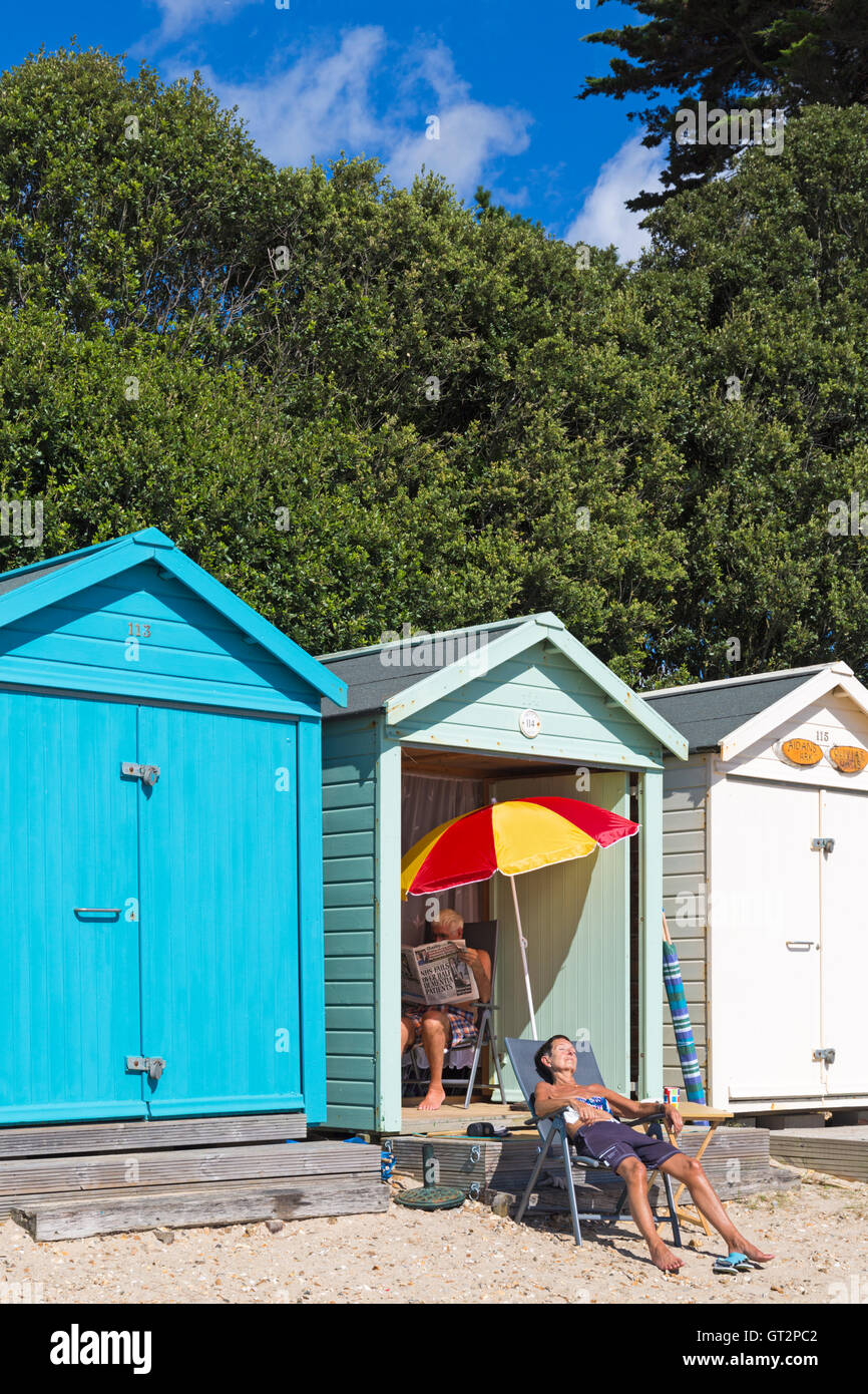 Senior woman sunbathing at beach huts with man sat inside in the shade ...