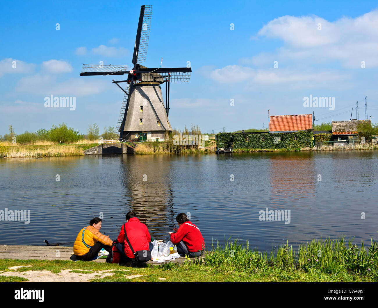 Three Chinese tourists picnicing at canal with a Dutch Windmill behind ...