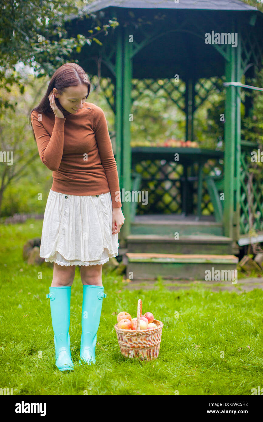 Young girl in rubber boots and basket with red apples at garden ...