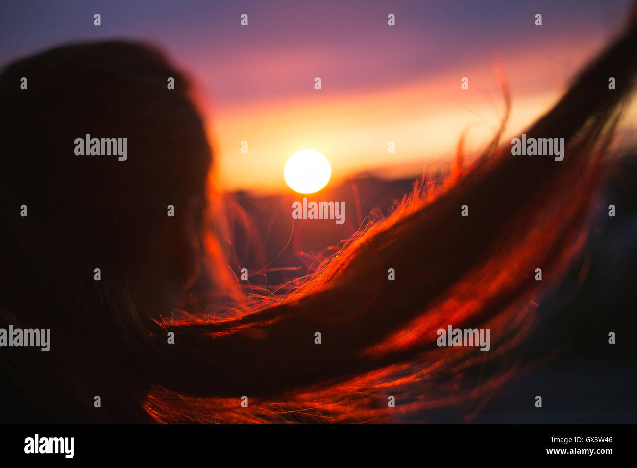 Back view of a beautiful young woman with sunset behind her brown hair ...