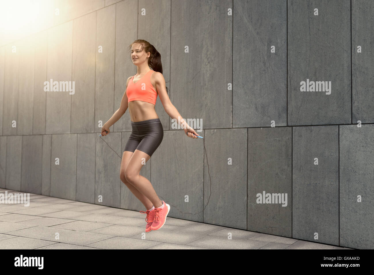 Fit smiling athletic young woman in shorts and top jumping rope outside ...