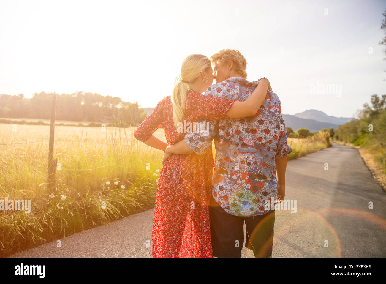 Rear view of romantic young couple strolling along sunlit rural road ...