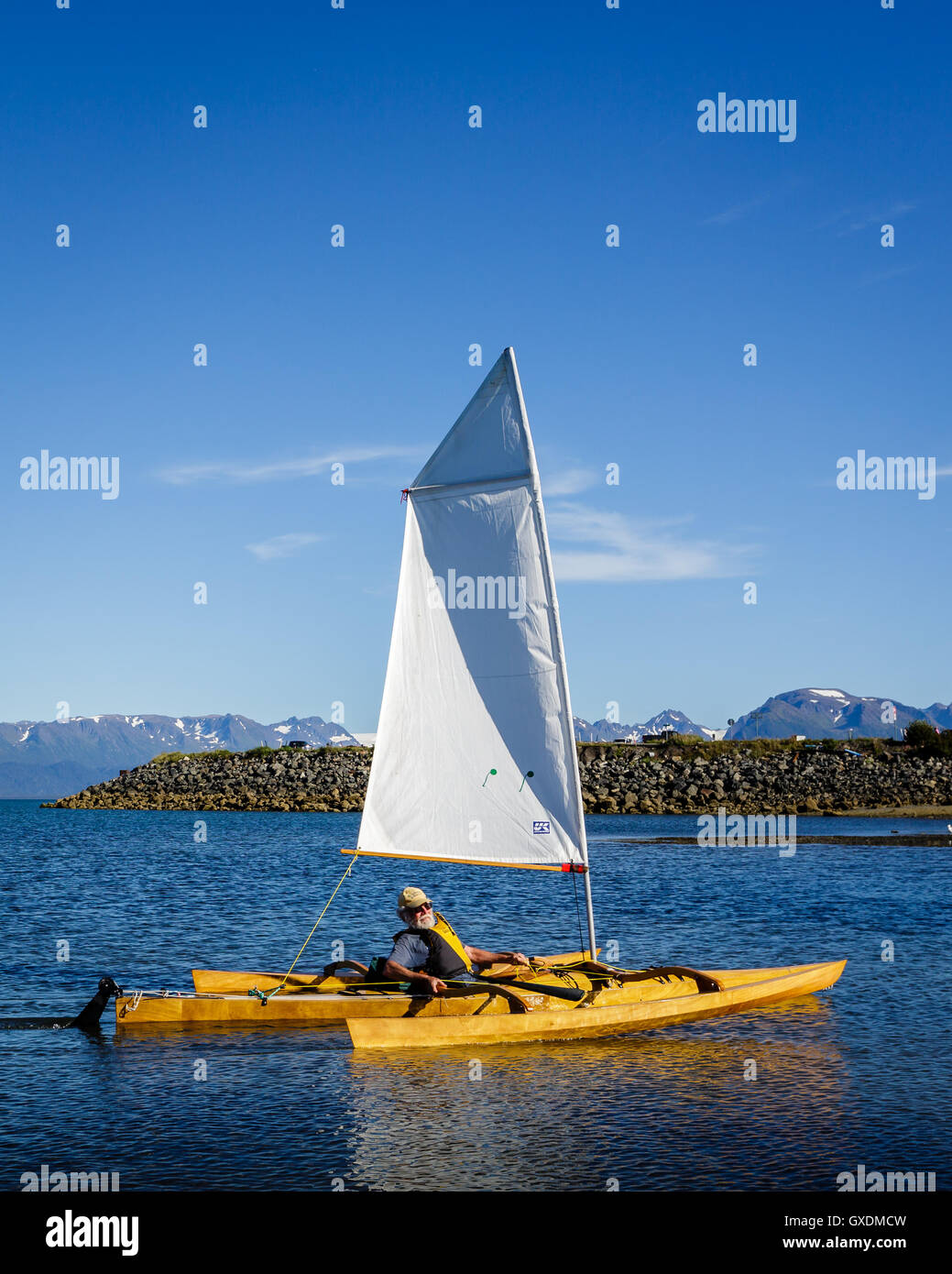 Man sails wooden outrigger sail boat past a breakwater on Homer spit ...