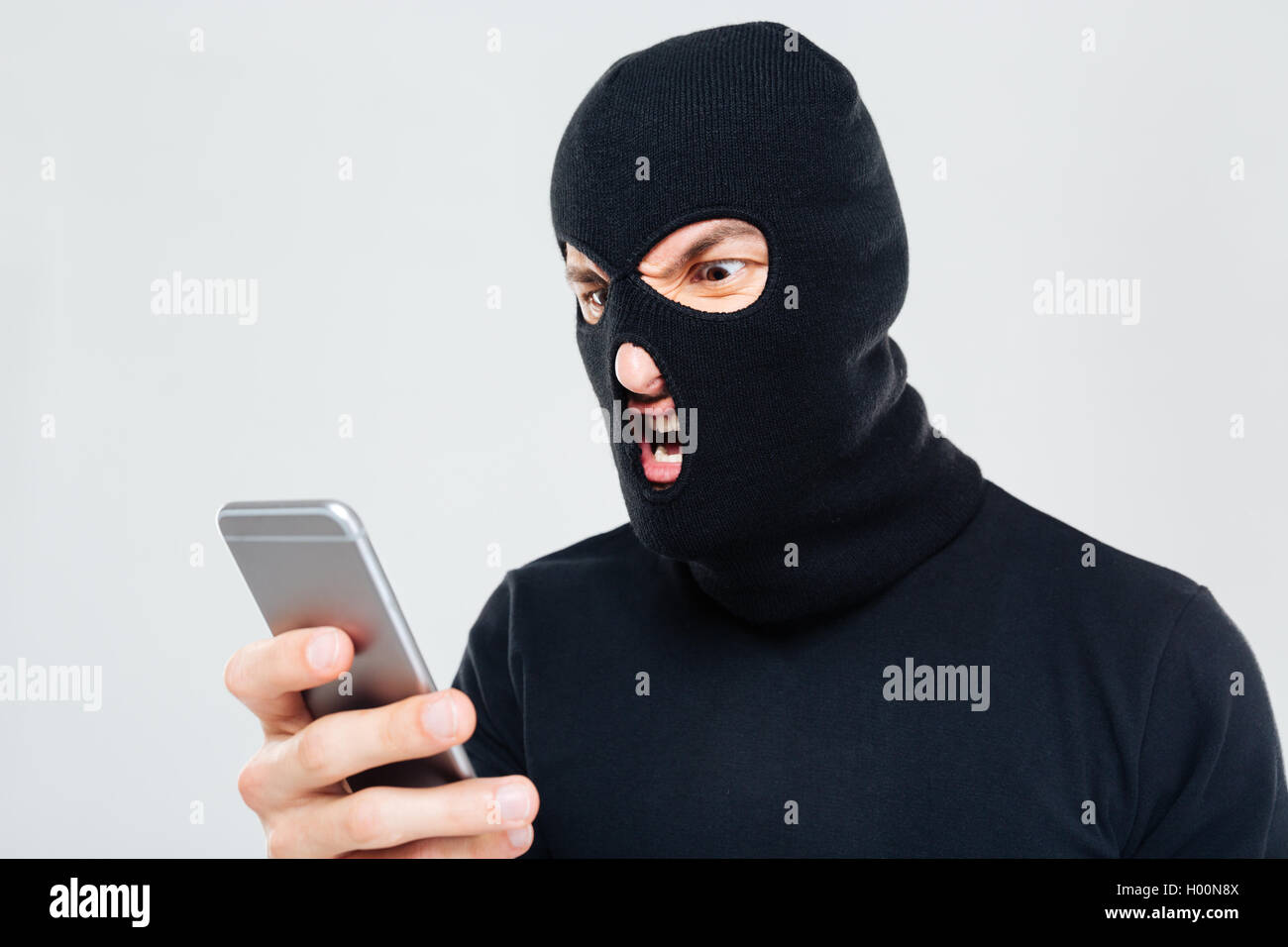 Mad aggressive young man in balaclava standing and using mobile phone ...