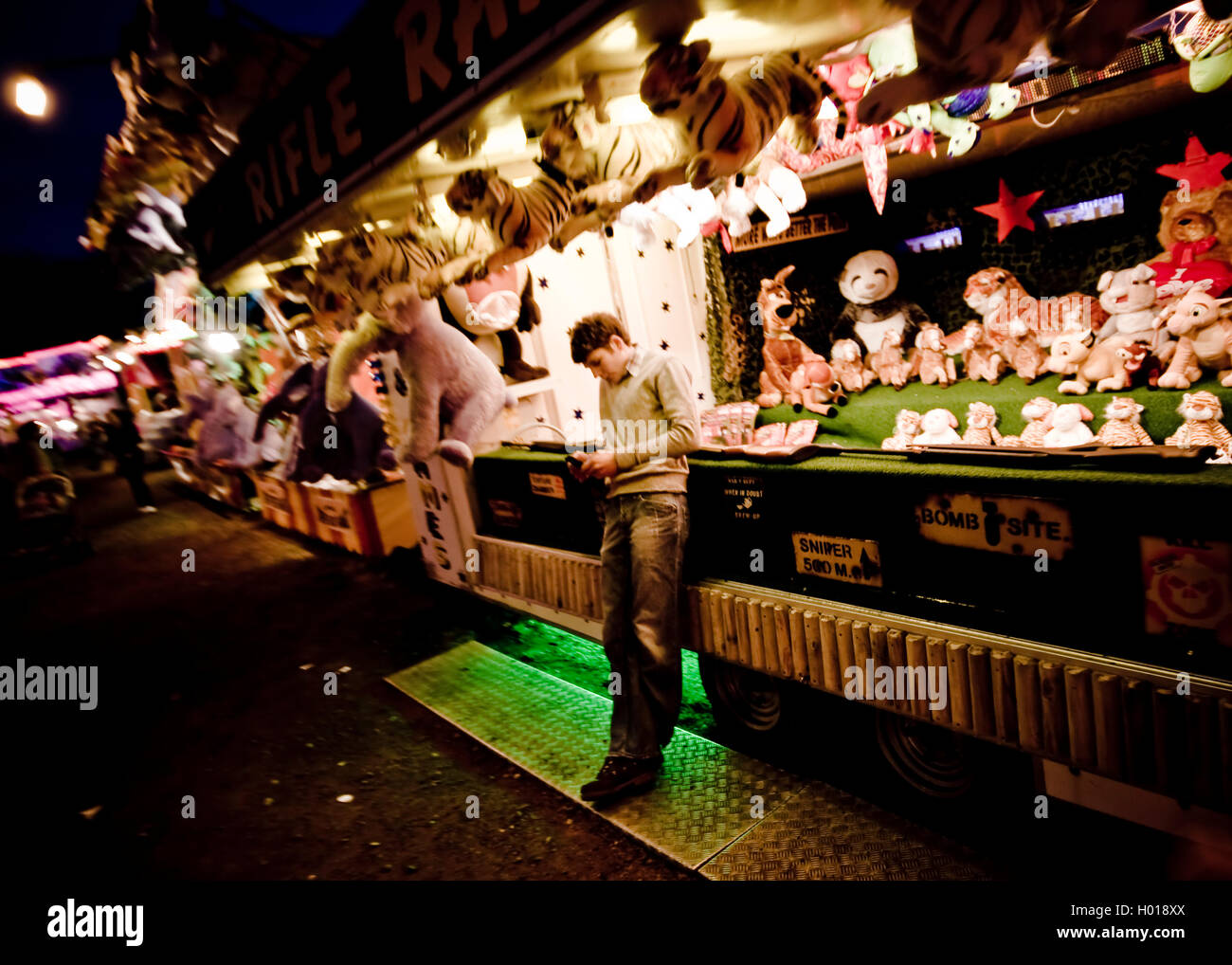 Young man looking at his phone in front of fairground stall at night ...