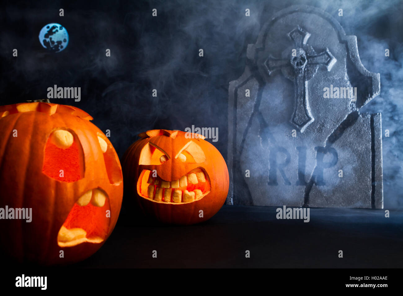 Angry face and scared face of Halloween pumpkins with moon and tombstone on misty dark ...