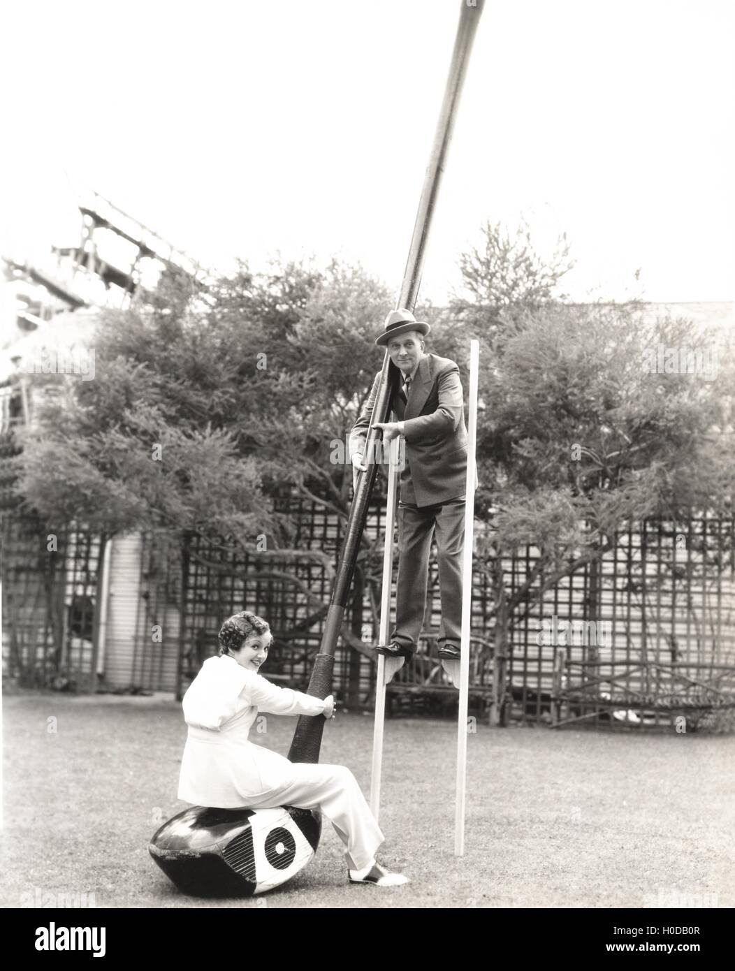 Man on stilts holding onto giant golf club with woman seated on it ...