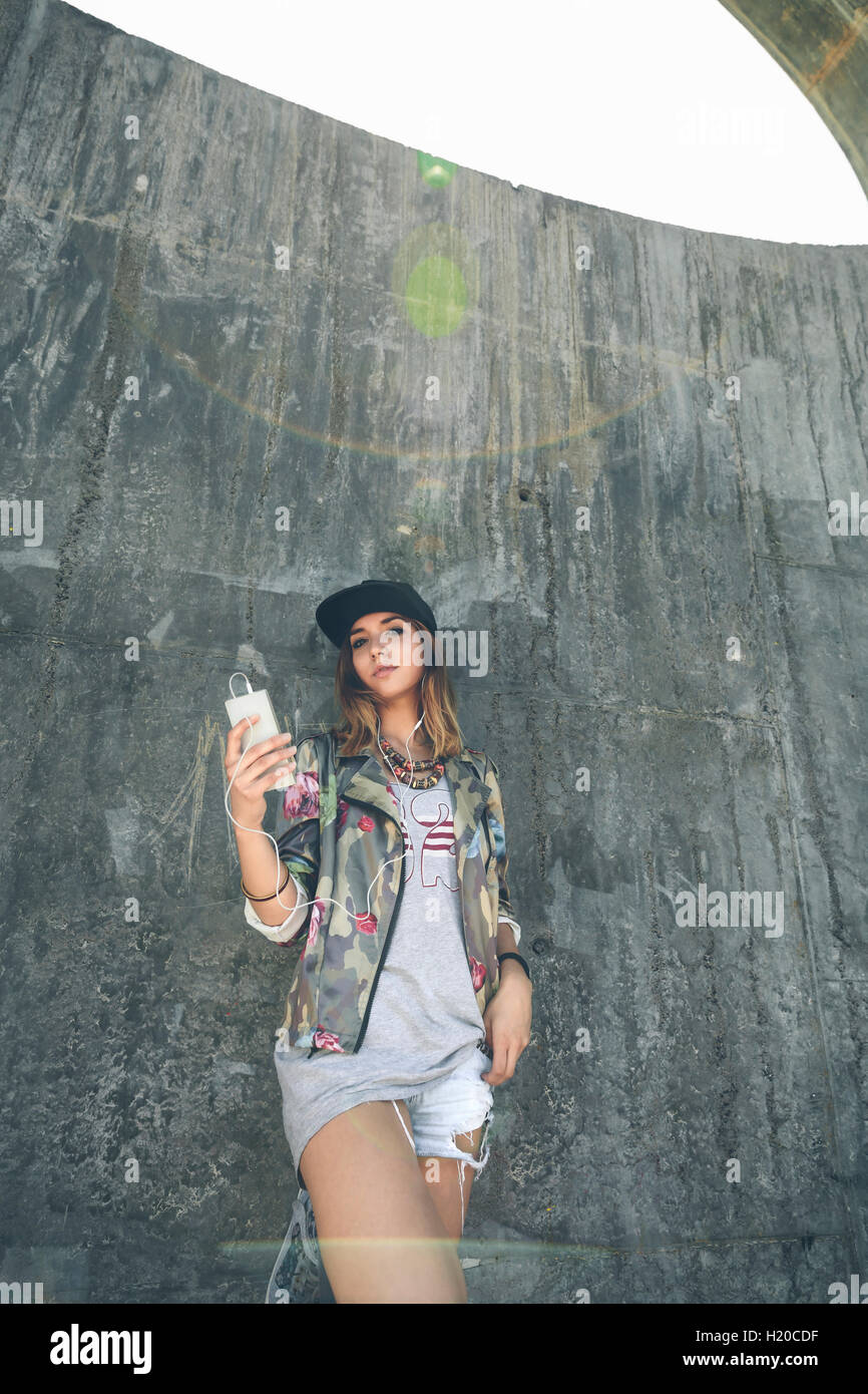 Young woman leaning against concrete wall listening music with ...