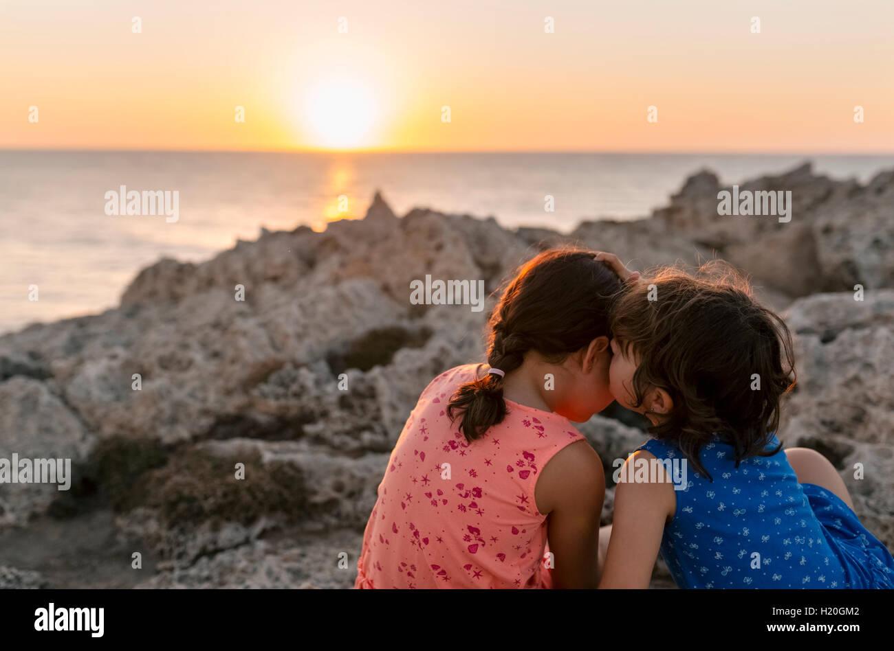 Back view of two little sisters sitting at rocky coast by sunset Stock ...