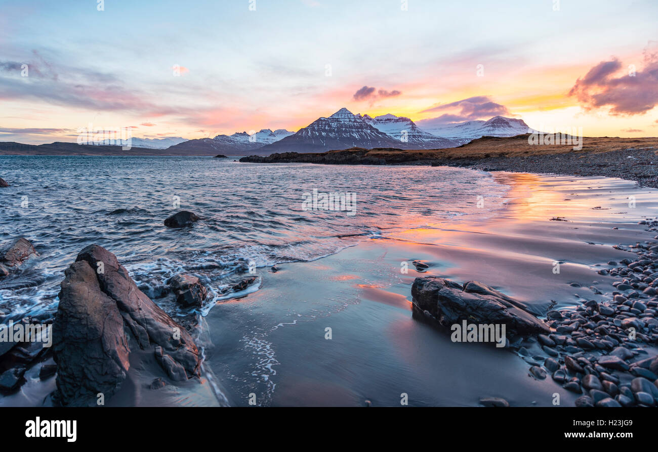 Sunset over snow-capped mountains, black beach in Berufjörður Fjord ...