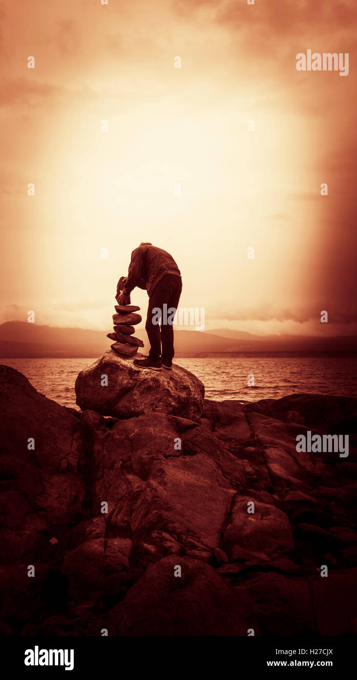 Man stacking rocks in a cairn at sunset by a lake processed with ...