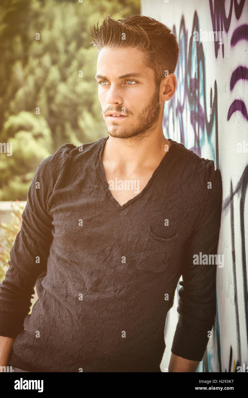 Attractive young man leaning against colorful graffiti wall, looking at ...