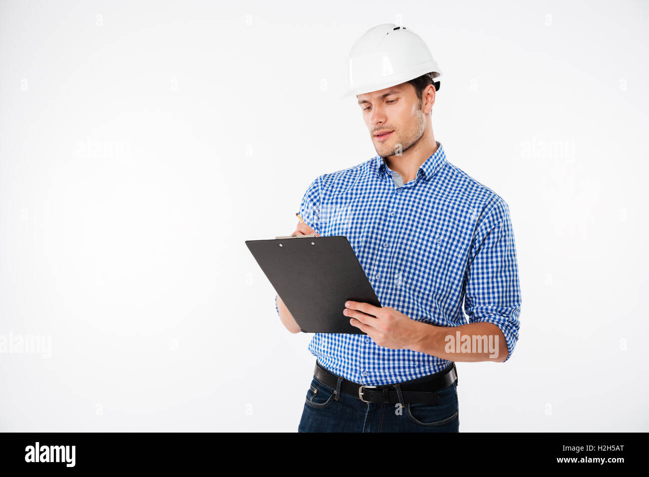 Concentrated young man builder in building helmet writing on clipboard ...
