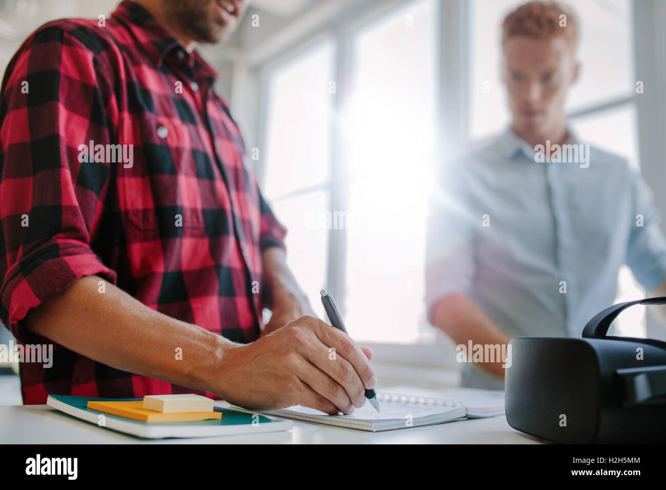 Closeup shot of young man writing on notepad, with colleague standing ...