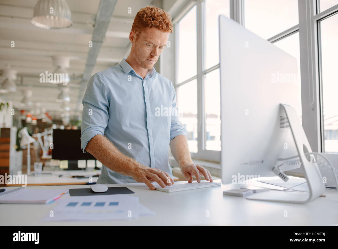 Shot of young man standing at his desk and working on computer ...