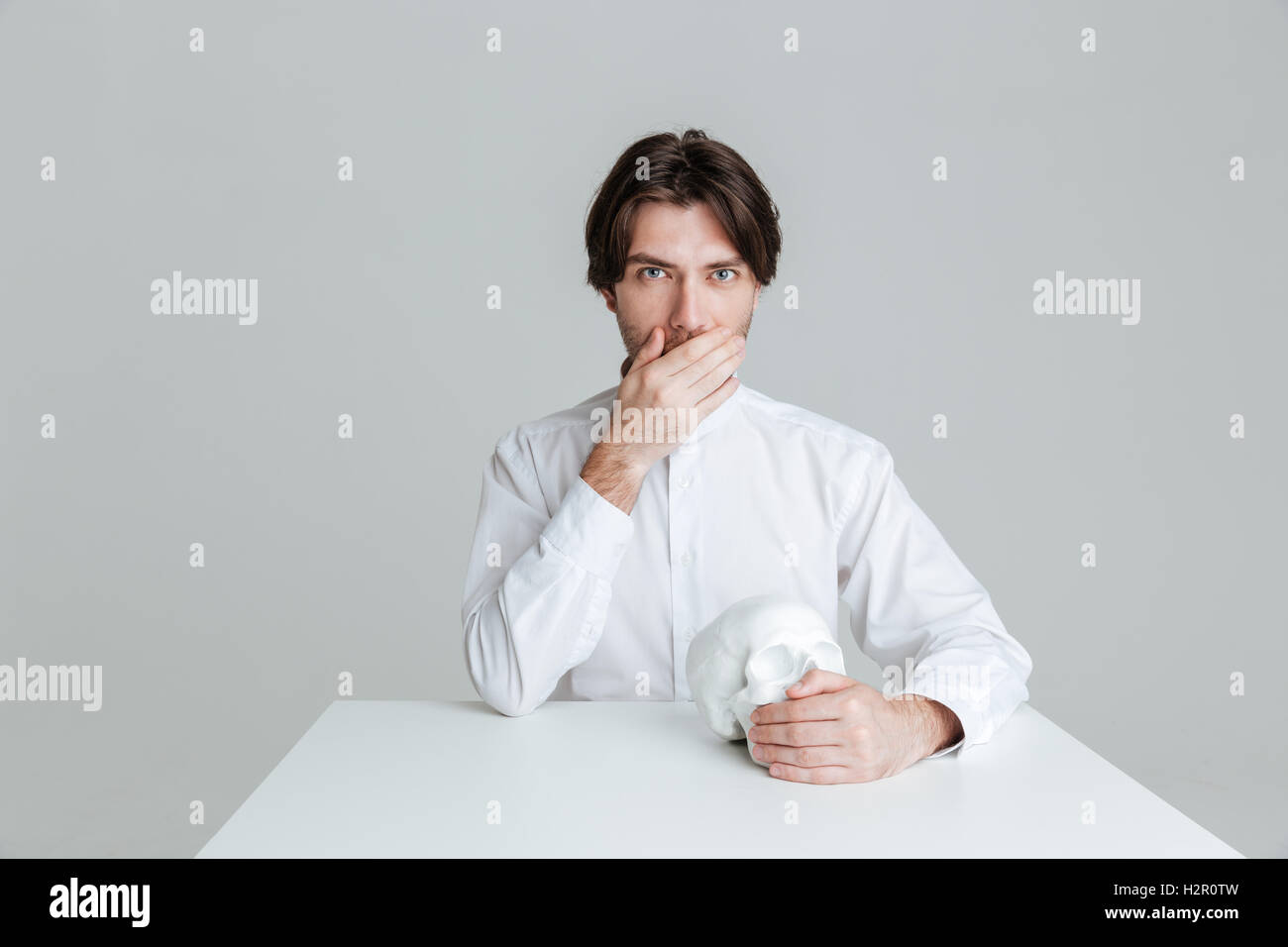 Pensive young man cover his mouth while sitting at the table and ...
