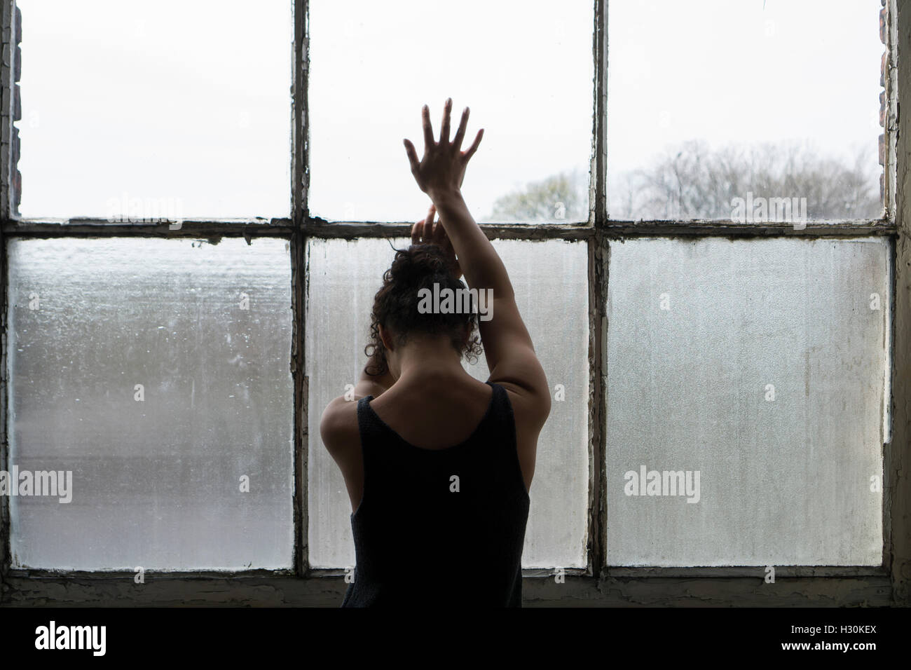 Rear view of a young woman standing by the window hands touching the ...