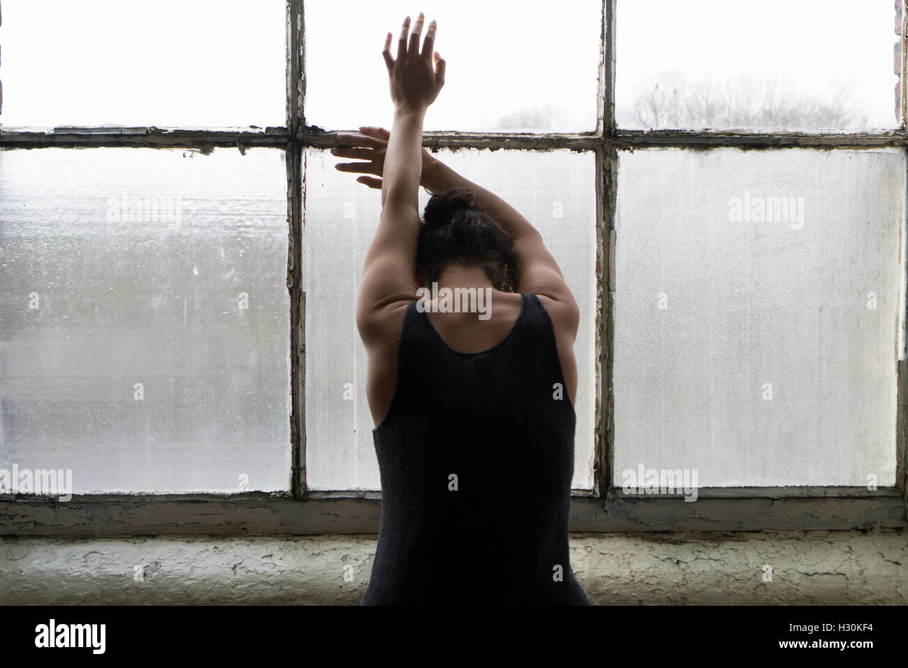 Rear view of a young woman standing by the window hands touching the ...