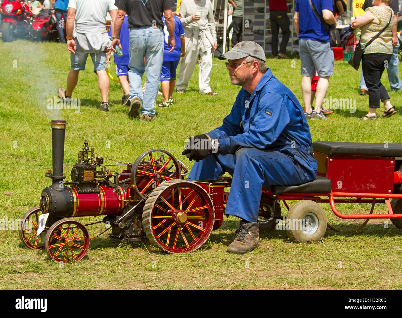 Man wearing blue overalls & cap seated on trailer towed by red scale ...