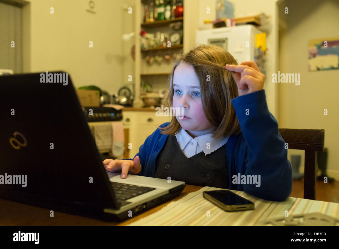 Little girl using a laptop computer at the kitchen table to do her ...