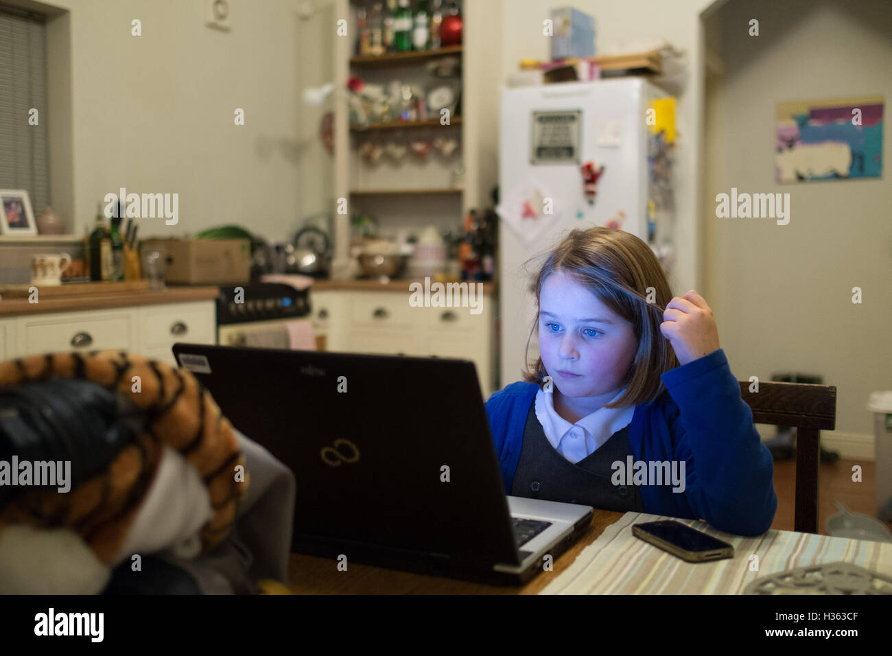 Little girl using a laptop computer at the kitchen table to do her ...