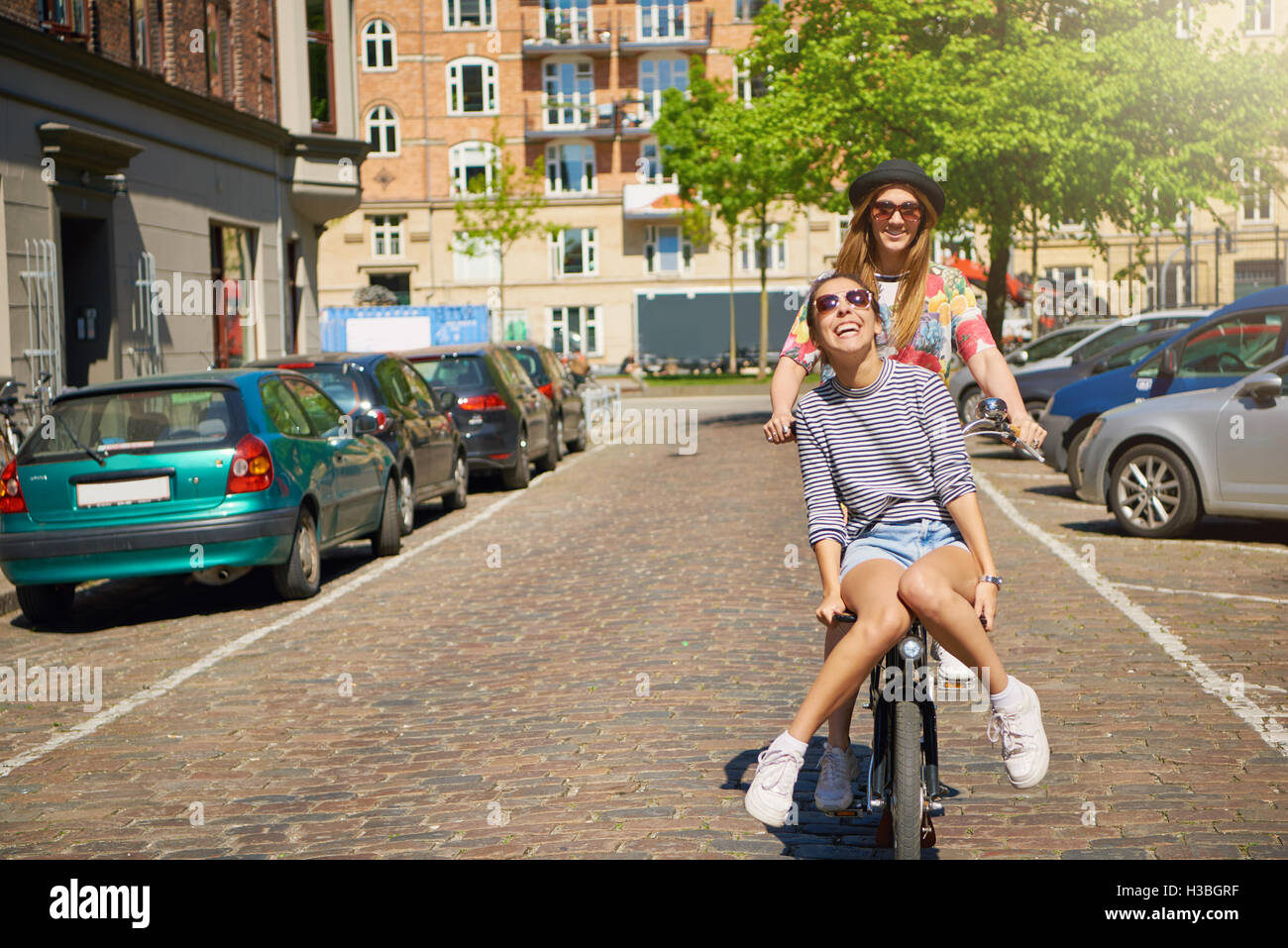Two young girlfriends having fun on a bicycle in a cobbled urban street ...