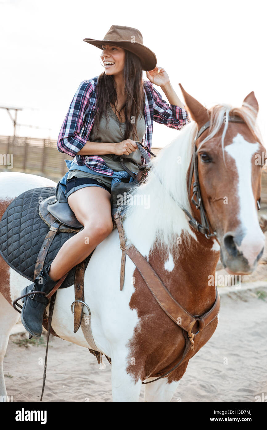 Cheerful pretty young woman cowgirl sitting and riding horse in village ...