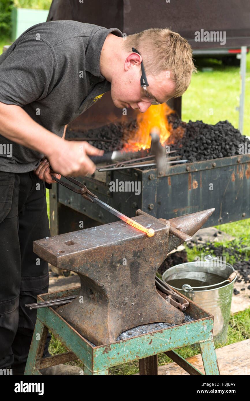 A blacksmith is hammering red hot metal on an anvil in front of a forge ...