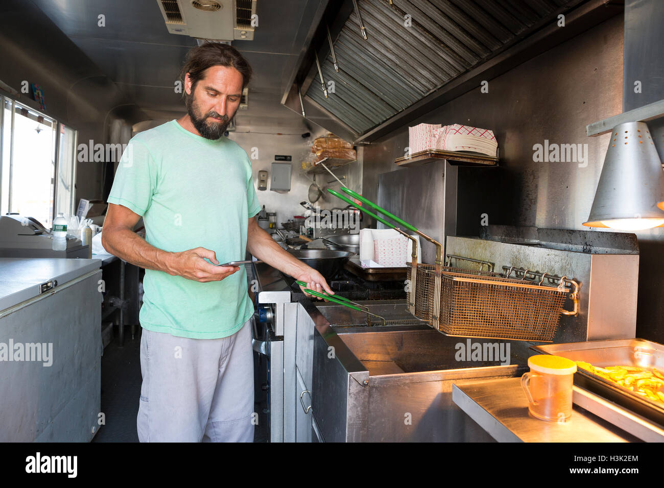 Man cooking food in fast food trailer whilst looking at smartphone ...