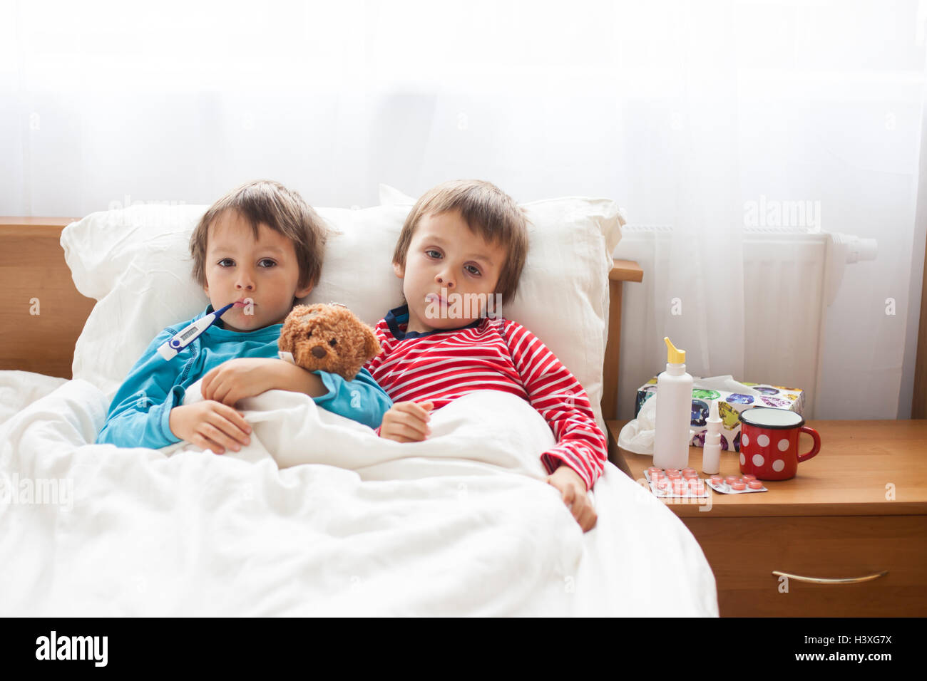 Two sick boys, brothers, lying down in bed with fever, holding teddy ...
