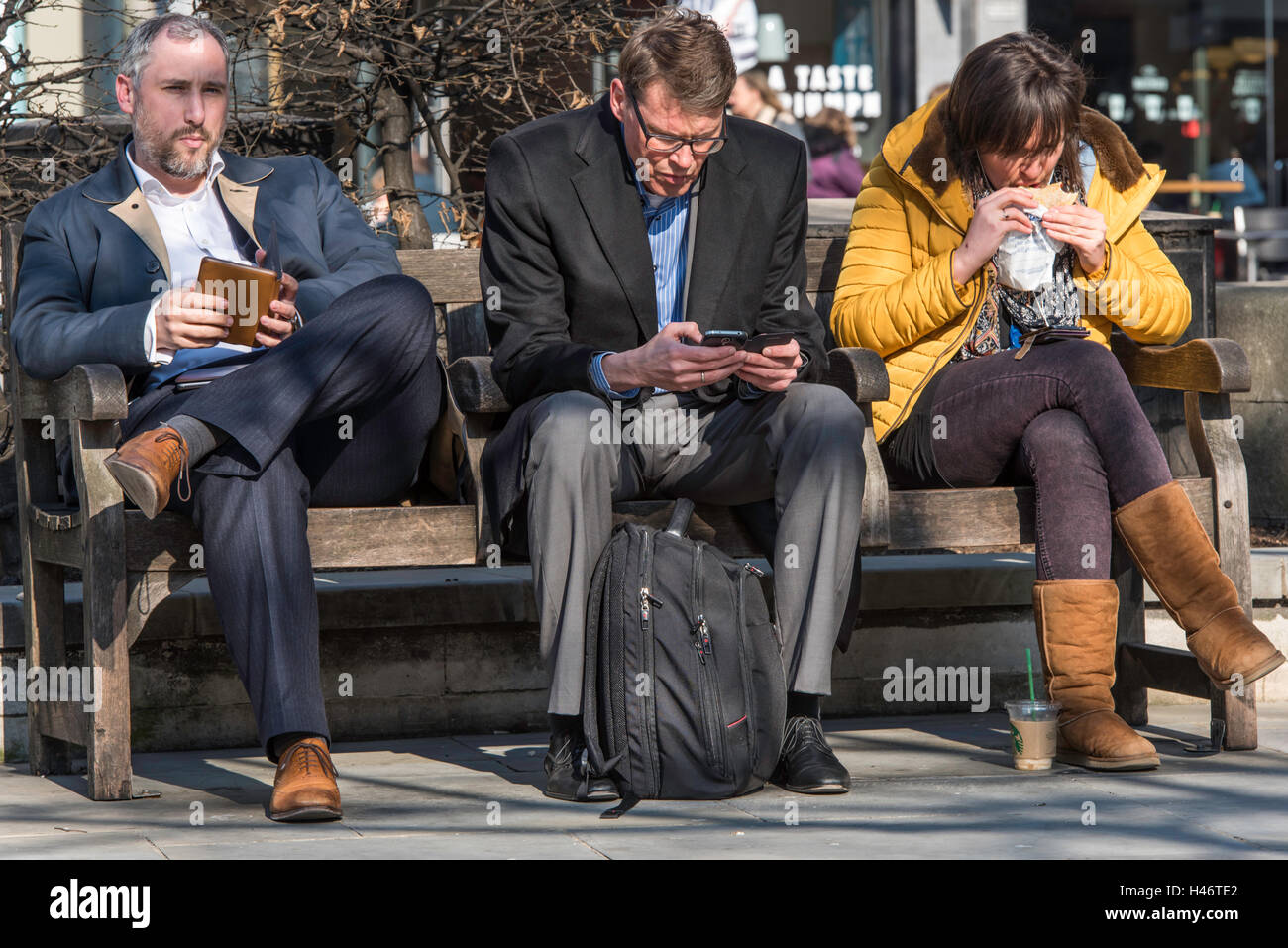 Three people sitting separately on a bench, eating and reading, during ...