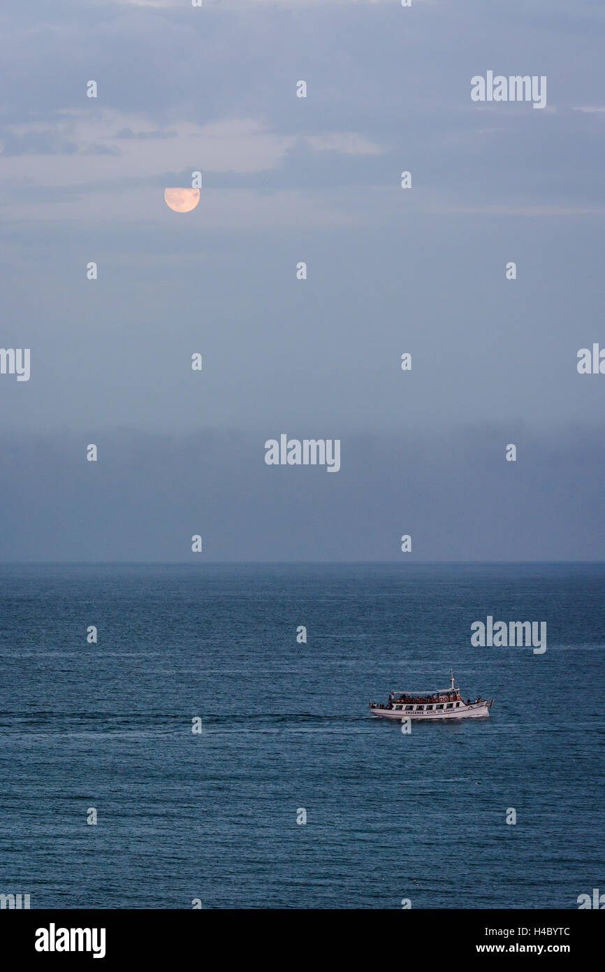 The full moon rises over the horizon as a pleasure boat passes in front ...