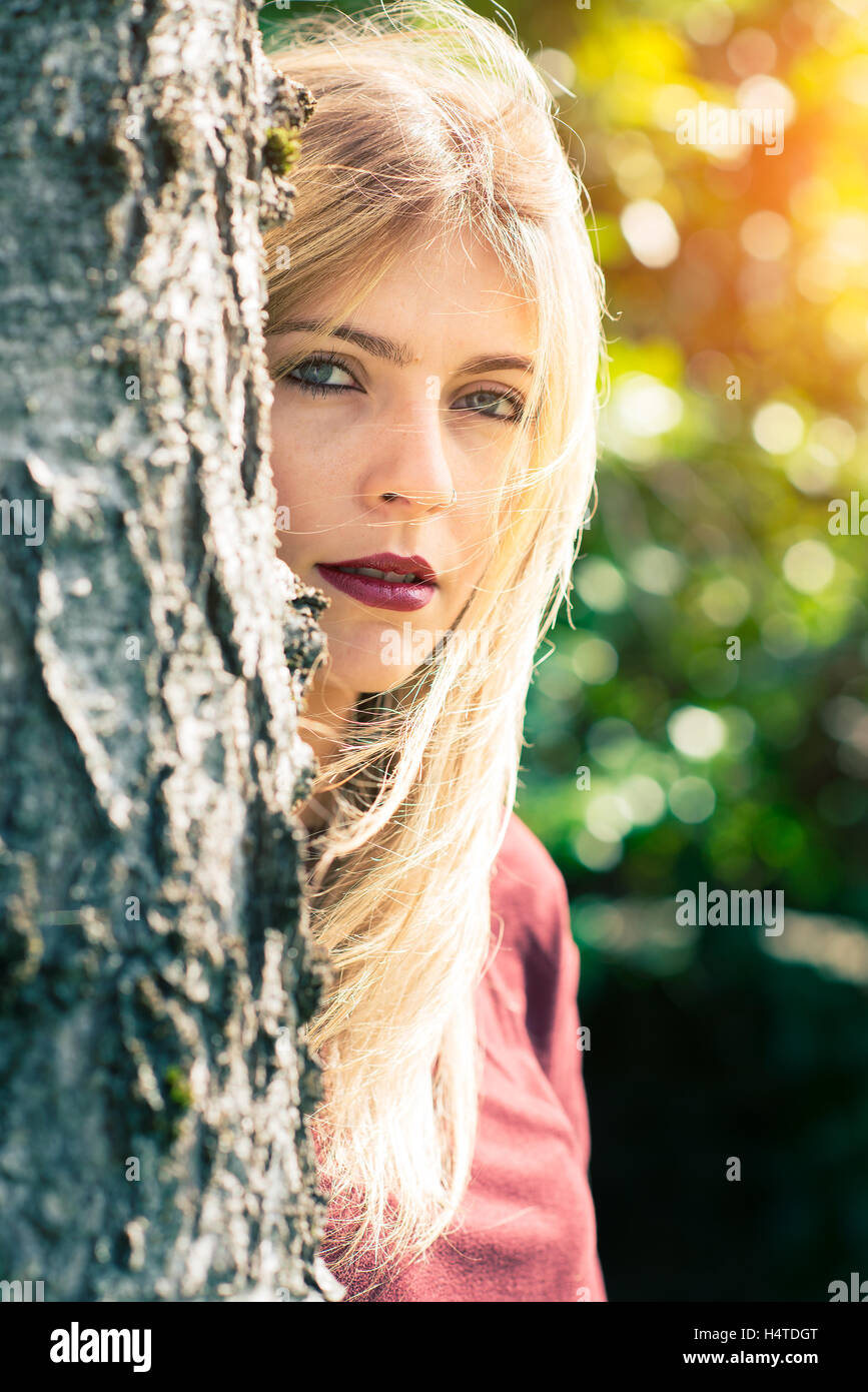 Blonde girl check and looks from behind a tree Stock Photo - Alamy