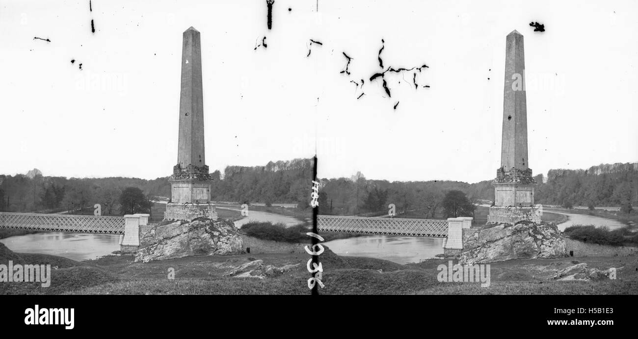 A square obelisk stands near a footbridge with an iron railing over a ...