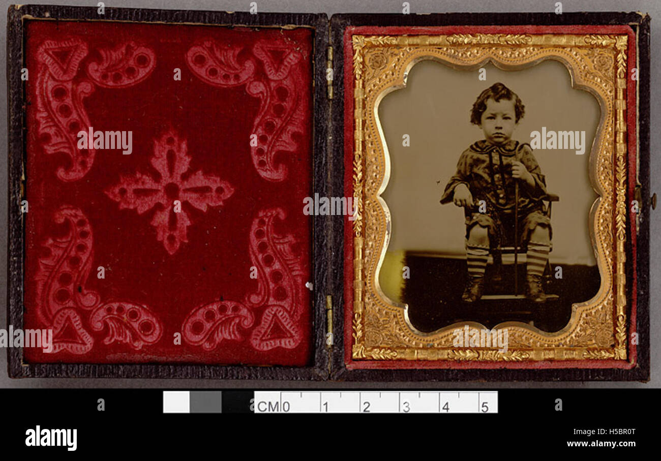 This photograph features a seated child holding a stick, offering a ...