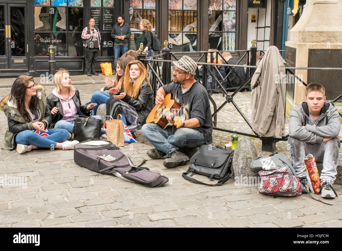 A group of teenagers sitting alongside a male busker who is playing his ...
