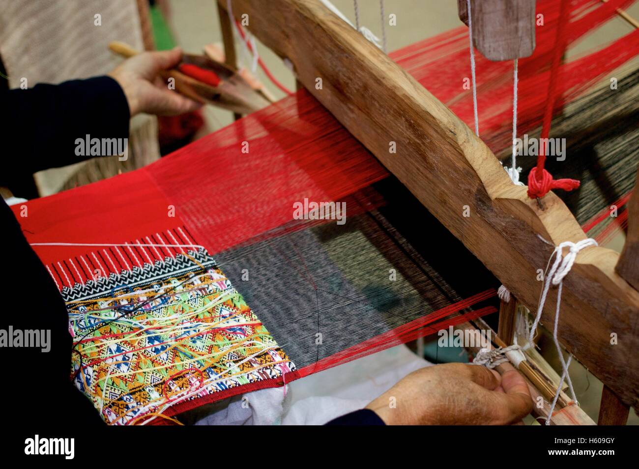 Old woman weaving red black and multi color cotton flag pattern on loom ...