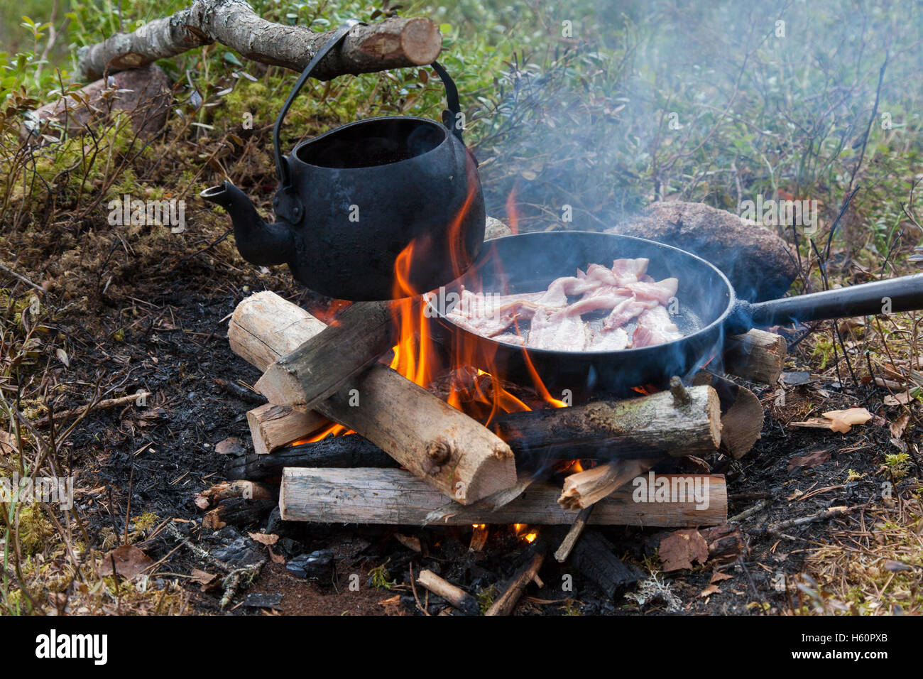 Blackened tin kettle boiling water and pan cooking bacon over flames ...