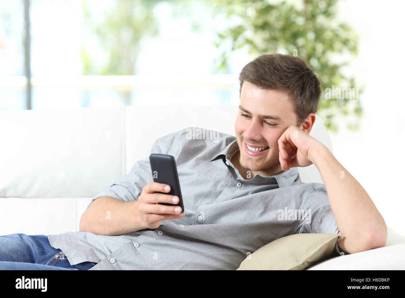 Happy man using a smartphone sitting on a couch at home with a window ...