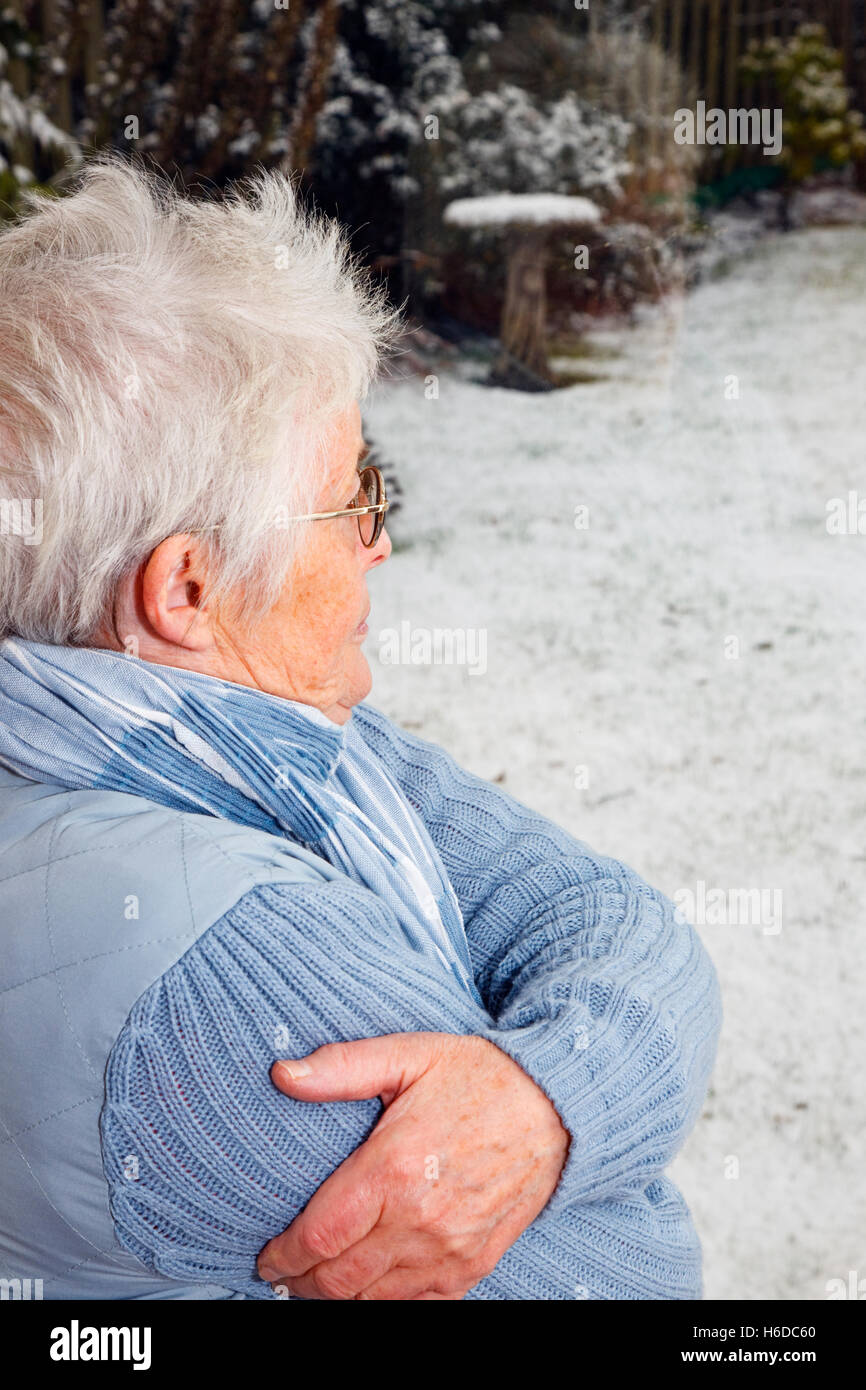 Senior woman feeling cold looking out of a window to snow in the garden ...